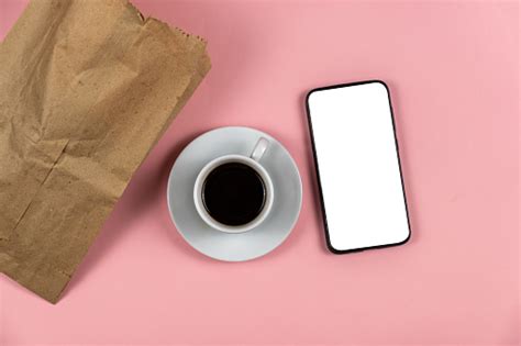 분홍색 배경에 커피 스마트 폰 및 종이 봉지 한 잔 접시가있는 풀 에스프레소 화이트 컵 샌드위치 햄버거 또는 점심 식사를 갈색 패키지로 포장하십시오 흰색 빈 화면이있는