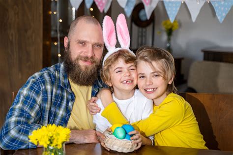 부활절 가족 전통 아빠와 토끼 귀를 가진 두 명의 백인 행복한 아이들은 함께 놀고 휴일을 위해 페인트로 계란을 장식하고 집 테이블에 함께 앉아 함께 즐거운 시간을 보냅니다