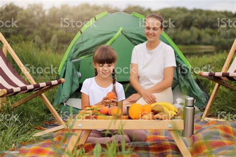 어머니와 그녀의 귀여운 딸이 강 근처에서 피크닉을하고 땅에 격자 무늬에 앉아 있고 카메라를보고 맛있는 음식과 신선한 공기를 즐기는 사람들의 초상화 2명에 대한 스톡 사진 및