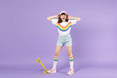 스튜디오에서 보라색 파스텔 배경에 고립 된 머리에 손을 넣어 노란색 스케이트 보드와 서 있는 생생한 옷에 흥분 십 대 소녀 사람들은 진심으로 감정 라이프 스타일 개념입니다