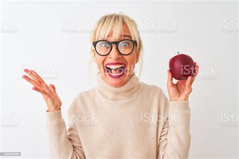 고립된 흰색 배경 위에 사과서를 들고 안경을 쓰고 있는 중년 여성은 매우 행복하고 흥분하며 큰 미소와 손을 들고 비명을 지르는 승리를 축하하는 우승자 표정 노인에 대한 스톡