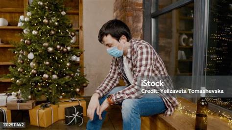 코비드 19 얼굴 마스크를 착용 외로운 남자는 매우 화가 술에 취해 창문과 크리스마스 트리에 앉아 병에서 술을 마시는 주제 알코올 중독 검역 코로나 바이러스 중 우울증