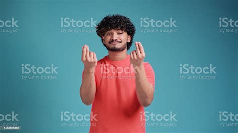 파란색 스튜디오 배경에 산호 티셔츠를 입고 수염을 기른 젊은 잘생긴 인도 남자가 손으로 돈 제스처를 하고 급여 지급을 요구한다 가리키기에 대한 스톡 사진 및 기타 이미지