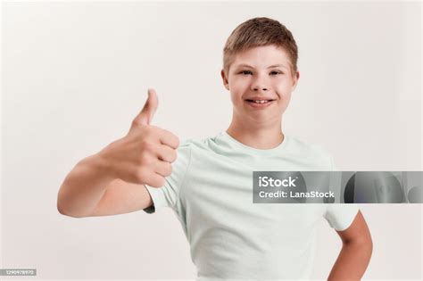 다운 증후군이 카메라에 미소 흰색 배경 위에 고립 된 서있는 동안 한 손으로 엄지 손가락을 보여주는 즐거운 장애인 소년 개발에 대한 스톡 사진 및 기타 이미지 Istock