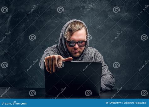 Programmer In Hoodie And Sunglasses Hacking Passwords On Laptop Sitting At A Desk In A Dark Room