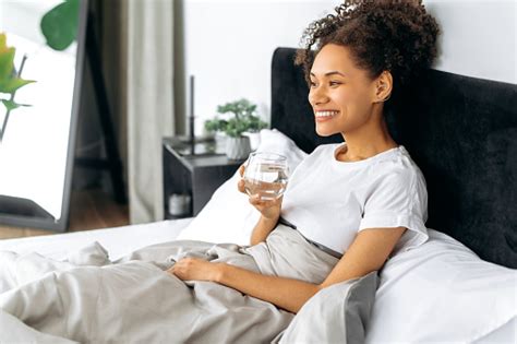 건강 관리 건강한 행복한 아프리카 계 미국인 곱슬 소녀는 물을 마시 며 하루를 시작하고 침대에 앉고 깨끗한 담수 한 잔을 들고 하루를 계획하고 신체의 물 속도를 돌 봅니다