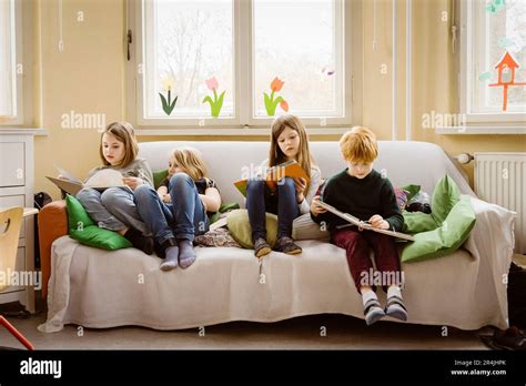 Full Length Of Girls And Boy Reading Books While Sitting On Sofa In