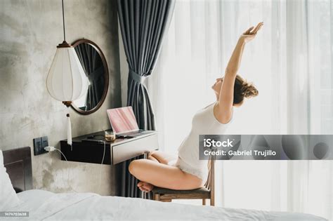 잠옷을 입은 젊은 여성의 전신 측면 모습은 아침 시간에 스트레칭을 하는 동안 음료와 컴퓨터 한 잔과 함께 테이블에 있는 의자에 다리를 꼬고 앉아 있다 45 49세에 대한