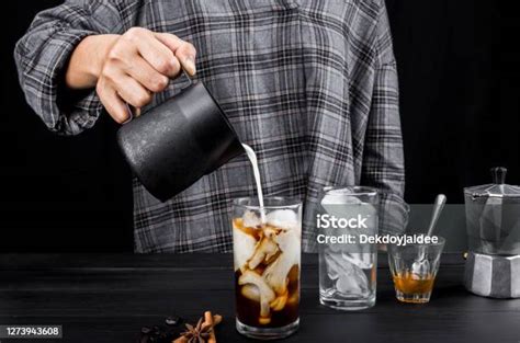우유를 붓는 타탄 셔츠를 입은 남자는 검은 색 배경 스튜디오에서 바 검은 나무 사진 촬영에 계피와 꿀을 곁들인 차가운 양조 아이스 커피를 만들고 모카 냄비 커피 강철에 대한