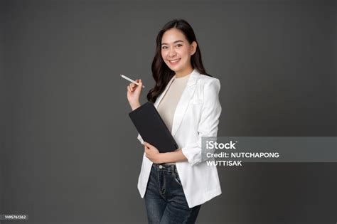 긴 머리를 한 아시아 여성 임원 미소 짓고 뛰어난 눈을 주고 태블릿과 펜으로 작업하고 흰색 양복을 입고 스튜디오에서 회색 장면으로 사진을 찍기 위해 서 있습니다 25 29