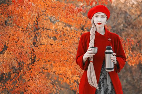 가을 배경에 빨간 코트에 긴 머리를 가진 금발 여자입니다 오렌지 단풍과 숲의 배경에 보온병 컵에서 차를 마시는 소녀 나뭇잎은 가지에서 떨어집니다 레드 터번과 세련된 코트