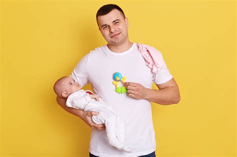 캐주얼 한 옷을 입고 미소 잘 생긴 남자는 손에 자신의 신생아와 노란색 벽에 서 카메라를 직접 보고 콩주머니를 들고 친자 휴가에 젊은 아빠 2 5 개월에 대한 스톡 사진 및