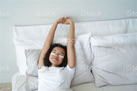 어린 소녀는 얼굴에 눈 밑 패치를 착용하고 깨어난 후 침대에서 스트레칭을 하고 있습니다 피부 관리 건강한 습관 20 24세에 대한 스톡 사진 및 기타 이미지 Istock