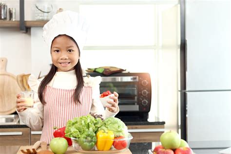 앞치마를 두르고 요리사 모자를 쓴 아시아의 웃는 소녀 아이의 초상화는 다채로운 신선한 야채 샐러드 음식 재료와 함께 부엌 카운터에 서 있고 귀여운 작은 요리사 아이가 건강한