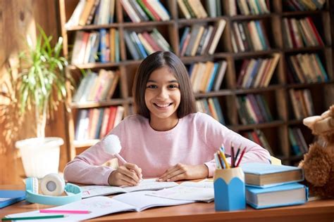 幸せなかわいいスマートなヒスパニック系インドのプレティーンの子供女子学生ラテン系の子供小学生生徒自宅のテーブルで勉強教室の机に座ってカメラを見