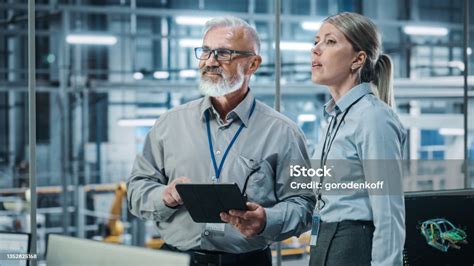 자동차 공장 여성 프로젝트 매니저가 태블릿 컴퓨터를 사용하여 남성 자동차 엔지니어와 계획 전략을 이야기합니다 전기 자동차 제조 자동화 된 로봇 암 조립 라인 생산 컨베이어