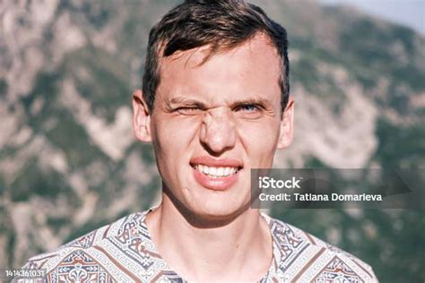 산을 배경으로 화창한 여름날 한쪽 눈을 가늘게 뜨고 카메라를 바라보며 웃고 있는 젊은 행복한 파란 눈동자 남성 밀레니얼 세대의 초상화 남자에 대한 스톡 사진 및 기타 이미지