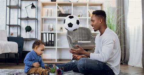 캐주얼 복장을 입은 행복한 흑인 아버지는 어린 아들과 함께 카펫에 앉아 공을 가지고 놀고 있습니다 엔터테인먼트 여가 시간 및 가족의 개념 사진 Unsplash의