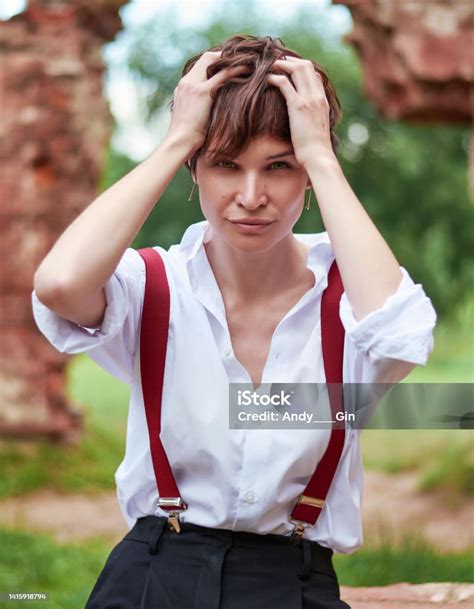 하얀 셔츠 검은 바지 붉은 색 서스펜더를 입은 세련된 아름다운 여성이 파괴 된 건물을 배경으로 공원에 앉아있는 이미지 스타일과 패션의 개념 30 34세에 대한 스톡 사진 및