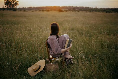 리넨 드레스를 입은 아름다운 여자가 소박한 의자에 앉아 여름 초원에서 일몰을 즐기고 있다 시골에서 책과 꽃 바구니로 휴식을 취하는 젊은 여성 분위기 있는 고요한 순간