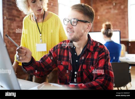 Young Smiling Analyst Pointing At Data On Computer Screen While Explaining It To Mature Employer