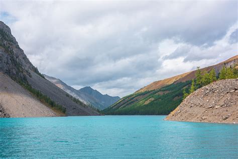 변화하는 날씨에 날카로운 바위 실루엣에 맞서 숲 언덕 사이에 큰 청록색 산 호수가있는 다채로운 가을 풍경 흐린 하늘 아래 햇볕이 잘 드는 계곡의 푸른 고산 호수에서 반짝이는