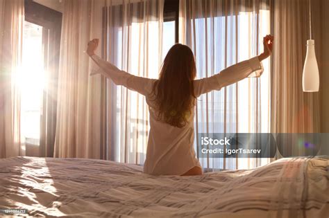 하얀 잠옷을 입은 사랑스러운 긴 머리의 여성은 아침에 침실에서 깨어나 아침 햇살에 침대에 앉아 팔을 옆으로 뻗습니다 일어나기에 대한 스톡 사진 및 기타 이미지 Istock