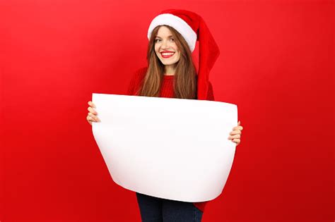 산타 모자를 쓴 매력적인 젊은 여성은 텍스트를 위한 공간이 있는 빈 흰색 포스터를 손에 들고 있습니다 빨간색 배경에 프로모터 25 29세에 대한 스톡 사진 및 기타 이미지