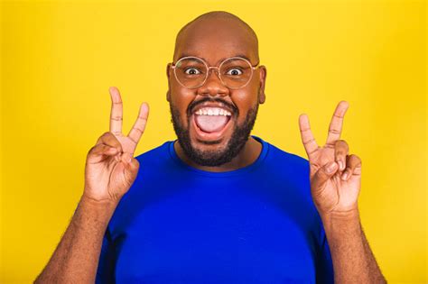 안경을 쓰고 노란색 바탕에 파란색 셔츠를 입은 잘생긴 아프리카 브라질 남자 손가락에 평화와 사랑의 표시 행복 기쁨 평화 사랑으로 미소 짓습니다 25 29세에 대한 스톡 사진