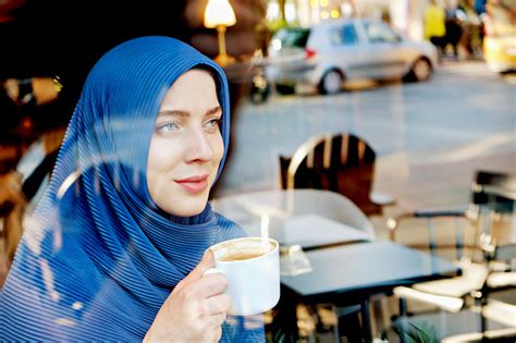 젊은 아름 다운 백인 여자 큰 전체 길이 창으로 소식통 커피 숍에서 전통적인 이슬람 머리 스카프를 입고 여자에 푸른 히 잡에 아늑한 카페 배경 복사 공간 초상화를 닫습니다