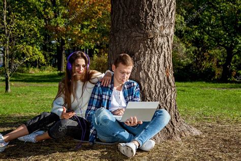 Молодая пара проводит время вместе в парке учится и работает сидя рядом с деревом с ноутбуком