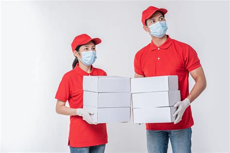 빨간 모자 빈 티셔츠 유니폼 얼굴 마스크 장갑을 낀 의식이 있는 젊은 배달원의 이미지는 밝은 회색 배경 스튜디오에 격리된 빈 흰색 판지 상자 팬데믹 코로나바이러스 바이러스