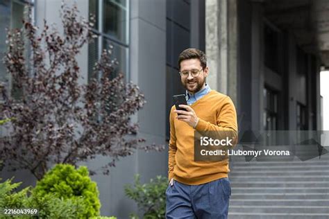 미소 짓고 성공한 젊은 남성 사업가가 사무실 건물 배경에 서서 바지 주머니에 손을 넣고 전화기를 사용하고 있습니다 30 34세에 대한 스톡 사진 및 기타 이미지 Istock