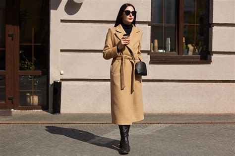 인기 레스토랑 앞에 서 있는 젊은 놀라운 여자의 초상화는 세련 된 선글라스 겨자 코트 가죽 가방 및 바지를 착용 합니다 여자는 거리에서 향기로운 커피와 함께 그녀의 시간을