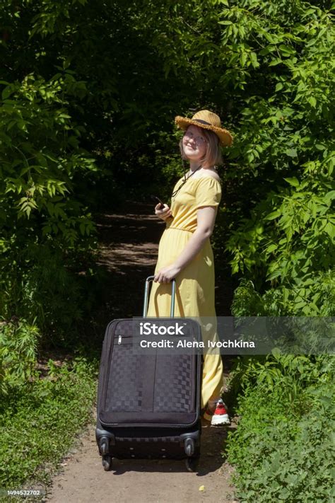밀짚모자를 쓰고 노란 드레스를 입은 아시아 소녀가 푸른 나무 사이로 길을 따라 커다란 검은색 여행 가방을 들고 있다 가방에 대한 스톡 사진 및 기타 이미지 Istock
