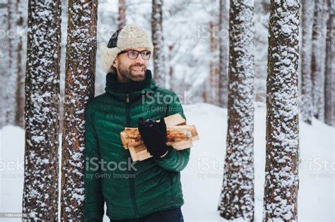 녹색 자 켓에서 행복 수염된 남자 나무 근처에 서 있다 명랑 식이 눈 덮인 숲 배경 스탠드 장작을 보유 하 고 신중 하 게 옆으로 보이는 야외 유행 잠겨있는 남성 40 44