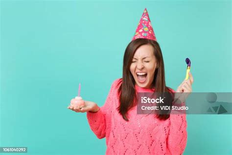 분홍색 스웨터에 미친 젊은 여자 연주 파이프 생일 모자 잡고 손에 케이크 촛불 눈 폐쇄 비명에 고립 된 파란색 배경 사람들의 라이프 스타일 개념입니다 복사 공간을 비웃는 다
