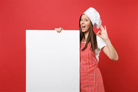스트라이프 앞치마 흰색 티셔츠 모자 요리사 모자 빨간 배경에 고립에서 주부 여성 요리사 요리사 베이커 여자 잡고 선전용 내용에 대 한 큰 백색 빈 게시판 복사 공간 개념을