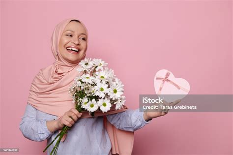 히잡을 쓴 쾌활한 무슬림 여성 꽃다발과 선물 상자를 들고 고립된 분홍색 배경을 옆으로 바라보며 웃고 있다 International Womens Day에 대한 스톡 사진 및