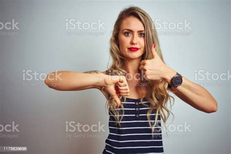 흰색 고립 된 배경 위에 서있는 줄무늬 티셔츠를 입은 젊은 아름다운 여성은 엄지 손가락을 위아래로 불일치와 동의 표현을하고 있습니다 미친 충돌 T 셔츠에 대한 스톡 사진 및