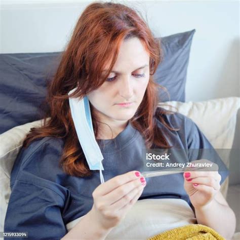 그녀의 얼굴에 마스크와 코로나 바이러스와 빨간 머리 여자는 온도계를 본다 의료 용 마스크에 집에서 침대에서 독감 바이러스를 가진 소녀 가정 생활에 대한 스톡 사진 및 기타