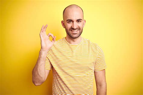노란색 고립 된 배경 에 캐주얼 줄무늬 티셔츠를 입고 수염을 가진 젊은 대머리 남자는 손과 손가락으로 확인 기호를하고 긍정적 인 미소 성공적인 식입니다 T 셔츠에 대한 스톡