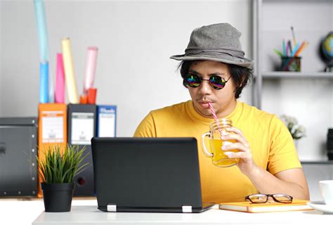 노트북을 사용 하 고 사무실에서 여름 휴가 시즌에 페도라 모자를 쓰고 오렌지 주스를 마시는 젊은이 개념에 대한 스톡 사진 및 기타 이미지 Istock