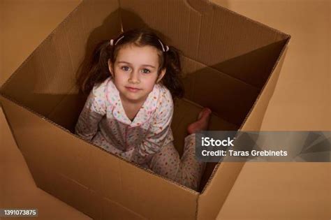 두 개의 포니 테일을 가진 매력적인 어린 유럽 소녀의 머리 위 전망 골판지 상자 안에 앉아있는 화려한 점이있는 잠옷을 입고 베이지 색 배경에 고립 된 카메라를보고있는 모습