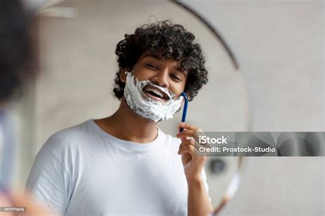 욕실에서 거울 근처에서 면도하는 인도 남자의 초상화 미용 루틴을 위해 면도기를 사용하여 얼굴에 면도 거품을 가진 남자 면도기에