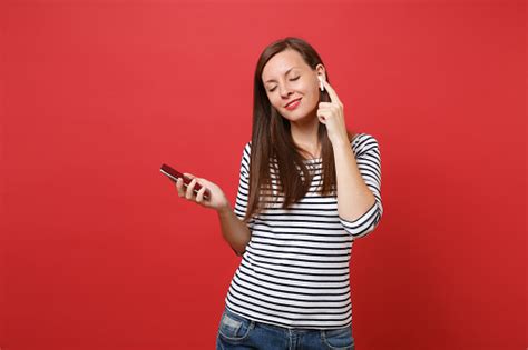 무선 이어폰 휴대폰을 들고 밝은 빨간색 배경에 고립 된 음악 듣기와 줄무늬 옷에서 편안한 젊은 여자 사람들이 진심으 감정 라이프 스타일 개념 복사 공간을 비웃는 다 갈색