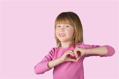 분홍색 배경에 분홍색 스웨터를 입은 어린 소녀의 초상화 아이는 행복한 표정으로 카메라를 향해 포즈를 취하며 손으로 심장 신호를 보여줍니다 스튜디오 촬영에 대한 스톡 사진 및