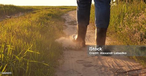 고무 장화를 신은 농부가 들판을 따라 먼지 투성이의 길을 따라 걷습니다 해질녘에 수확한 들판을 밟는 남자의 발 먼지를 일으키고
