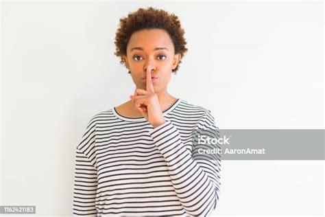 젊은 아름 다운 아프리카 계 미국인 여자 흰색 배경에 줄무늬 스웨터를 입고 입술에 손가락으로 조용 하 게 요구 침묵과 비밀 개념
