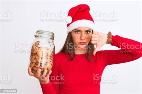 화가 얼굴 아래로 엄지 손가락으로 싫어하는 것을 보여주는 부정적인 기호 거부 개념으로 고립 된 흰색 배경 위에 쿠키의 항아리를 들고 크리스마스 산타 모자를 입고 아름다운